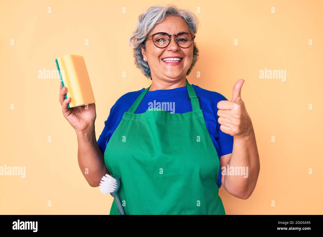 Senior hispanic grey- haired woman wearing apron holding scourer ...