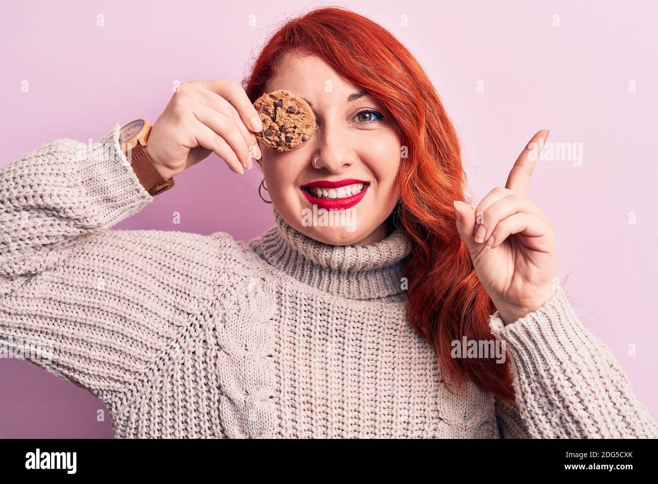 Young beautiful redhead woman standing holding chocolate cookie over ...