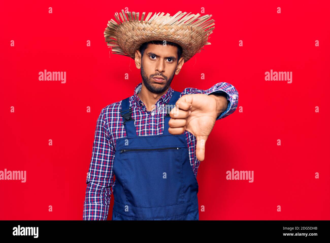 Young latin man wearing farmer hat and apron looking unhappy and angry ...