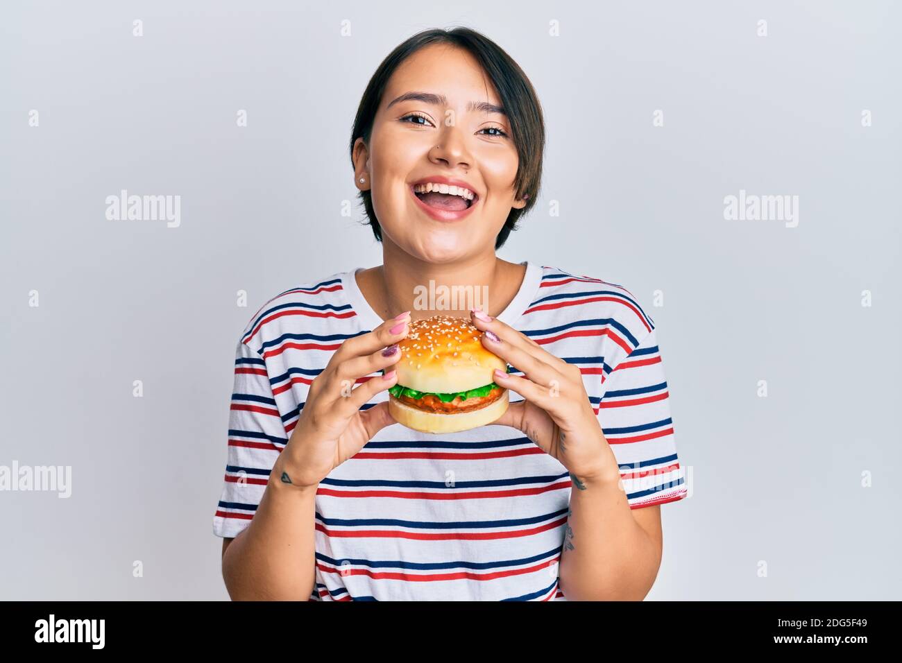 Beautiful young woman with short hair eating hamburger smiling and ...