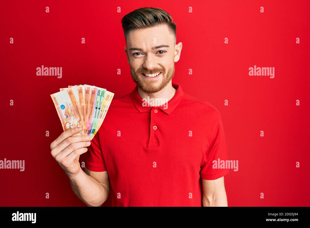 Young redhead man holding philippine peso banknotes looking positive ...