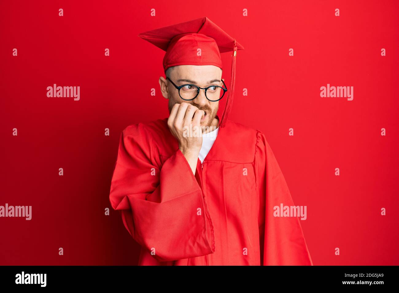 Young redhead man wearing red graduation cap and ceremony robe looking ...