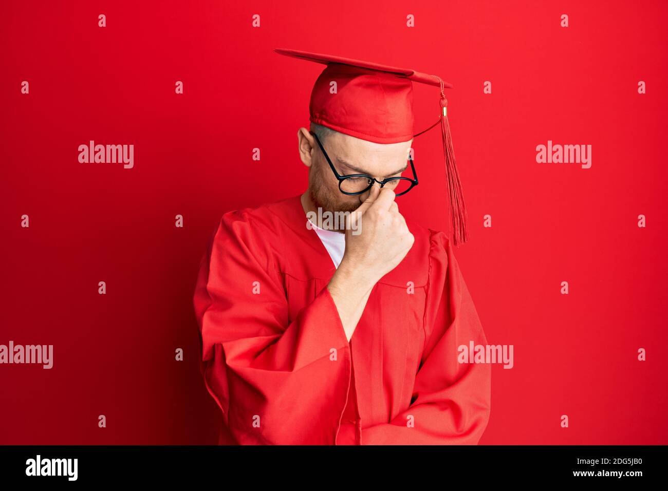 Young redhead man wearing red graduation cap and ceremony robe tired ...