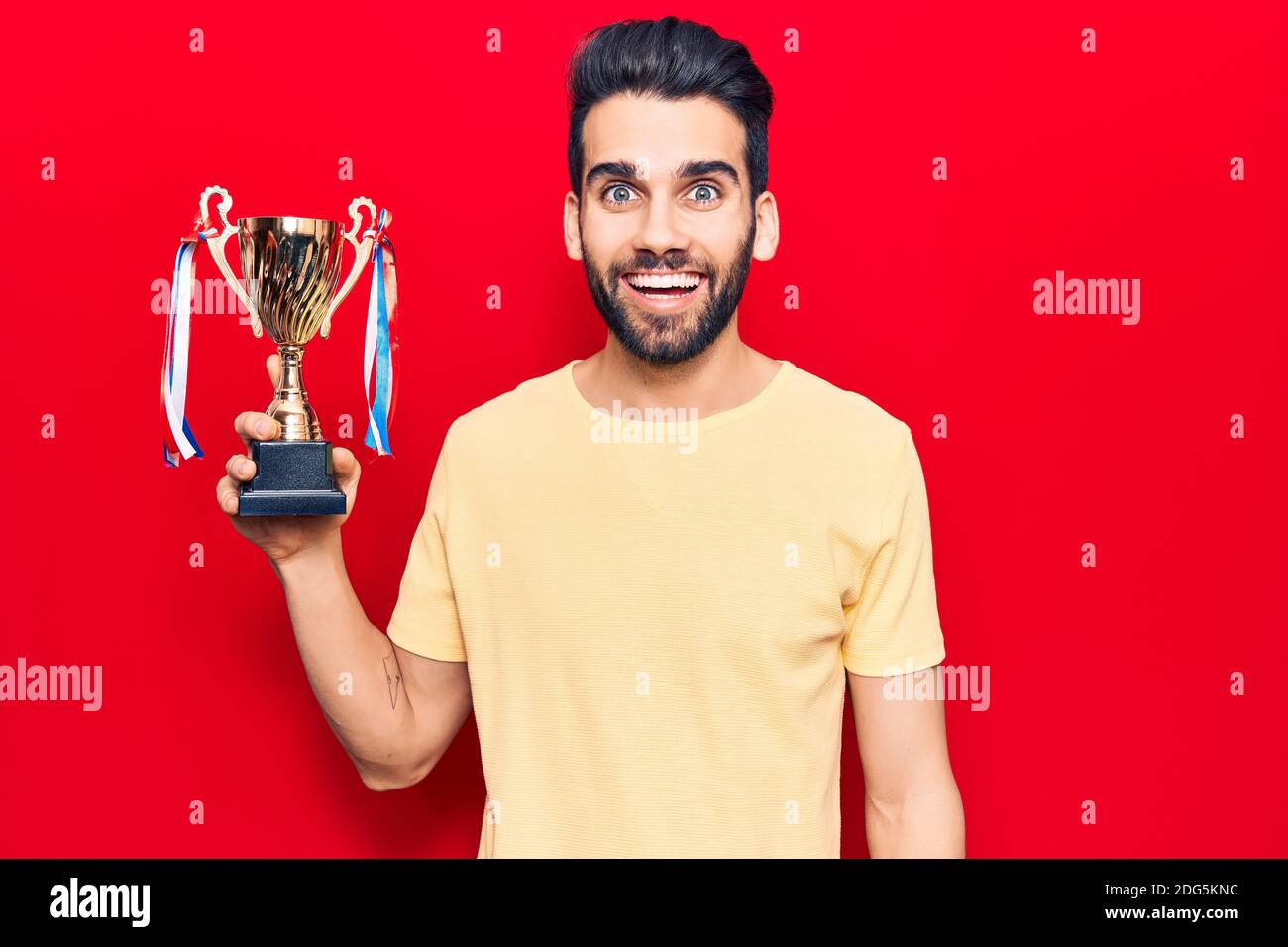 Young handsome man with beard holding trophy looking positive and happy ...