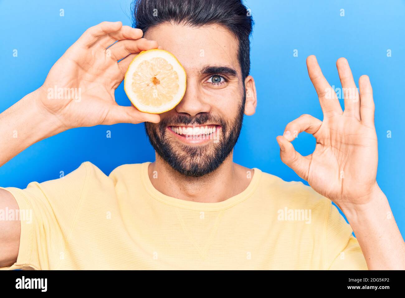 Young handsome man with beard holding slice of lemon over eye doing ok ...