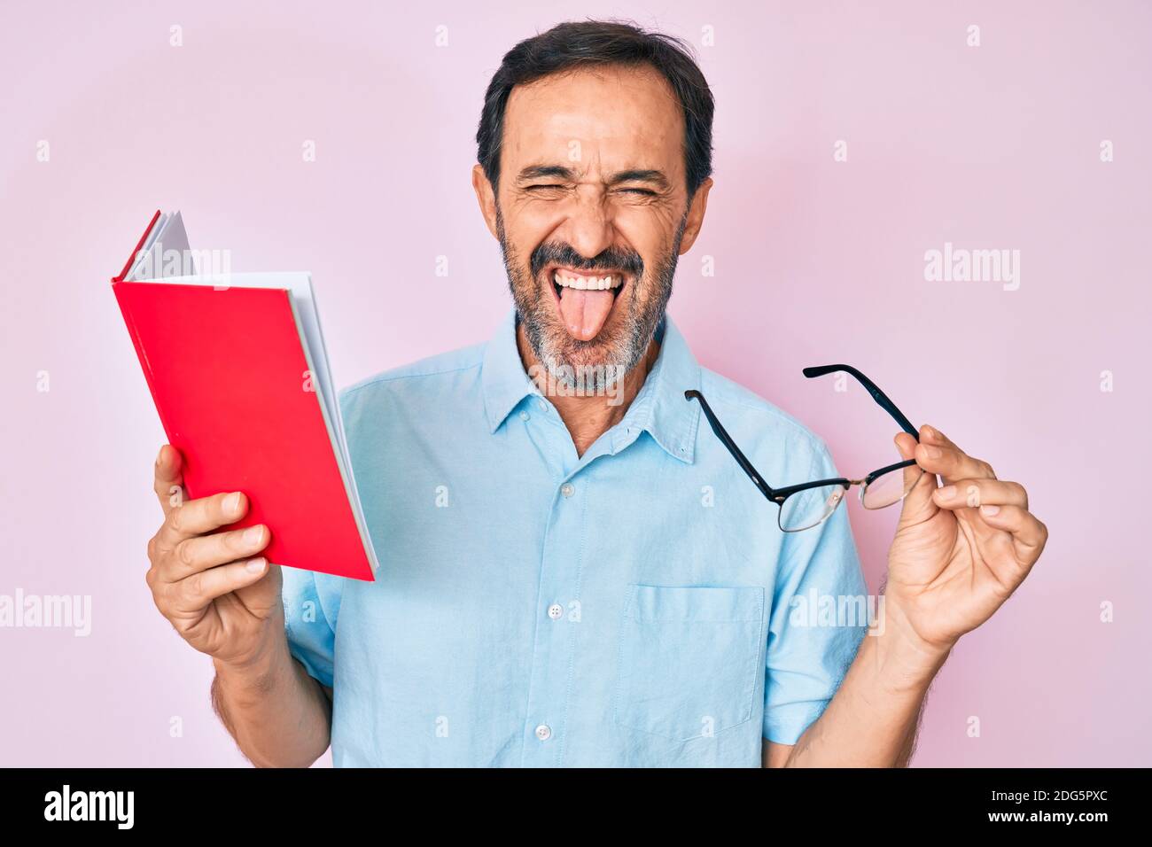 Middle age hispanic man reading book holding glasses sticking tongue ...