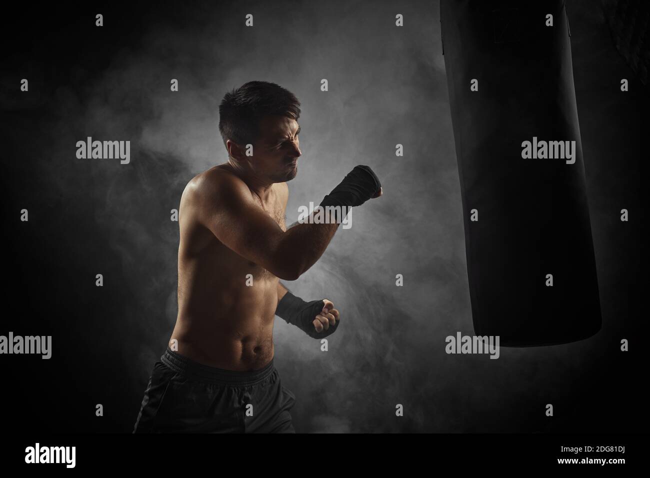 Aggressive boxer in black boxing wraps punching in boxing bag on dark ...