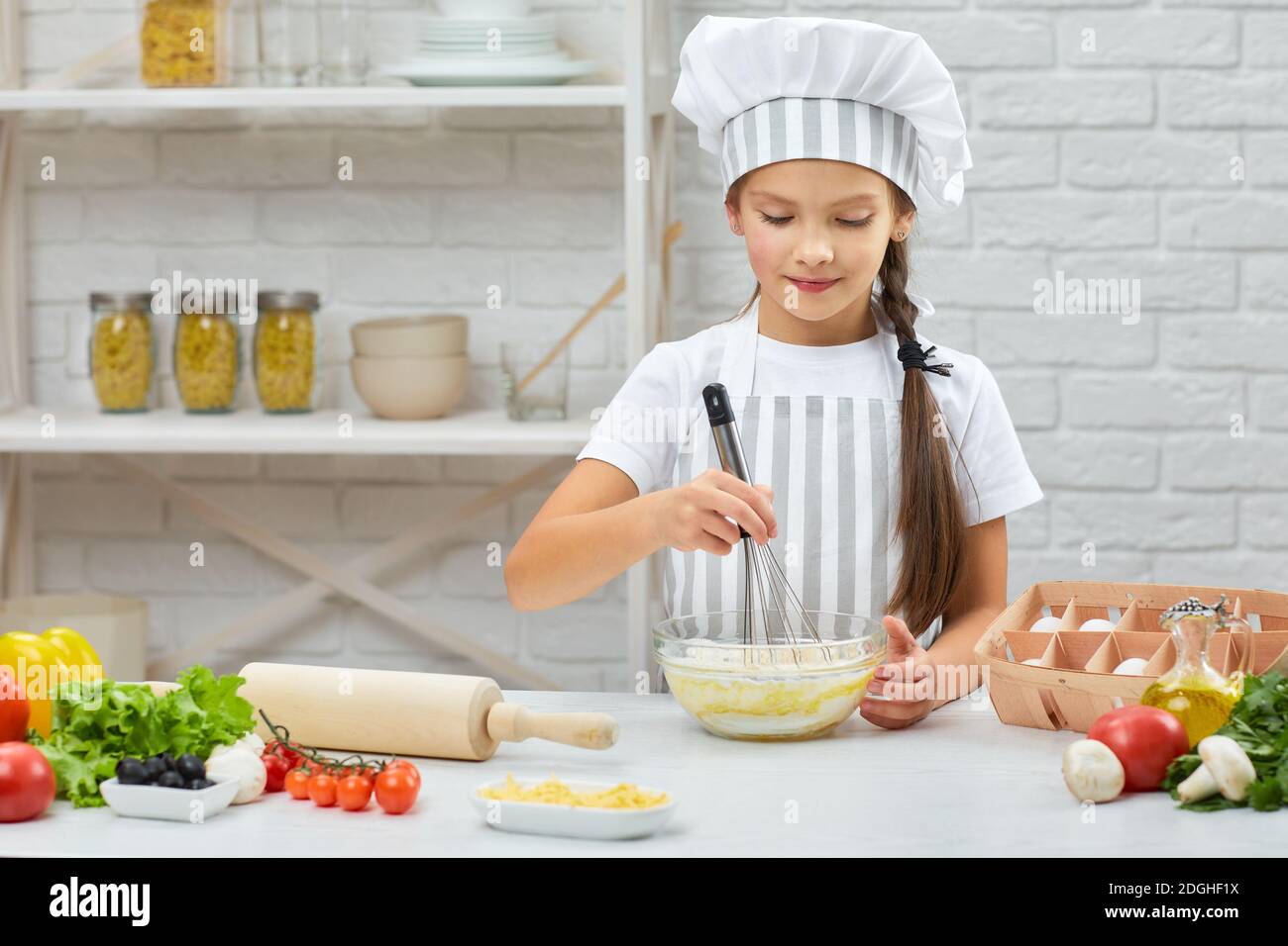 little child girl in cap and an apron kneads with steel whisk the dough ...