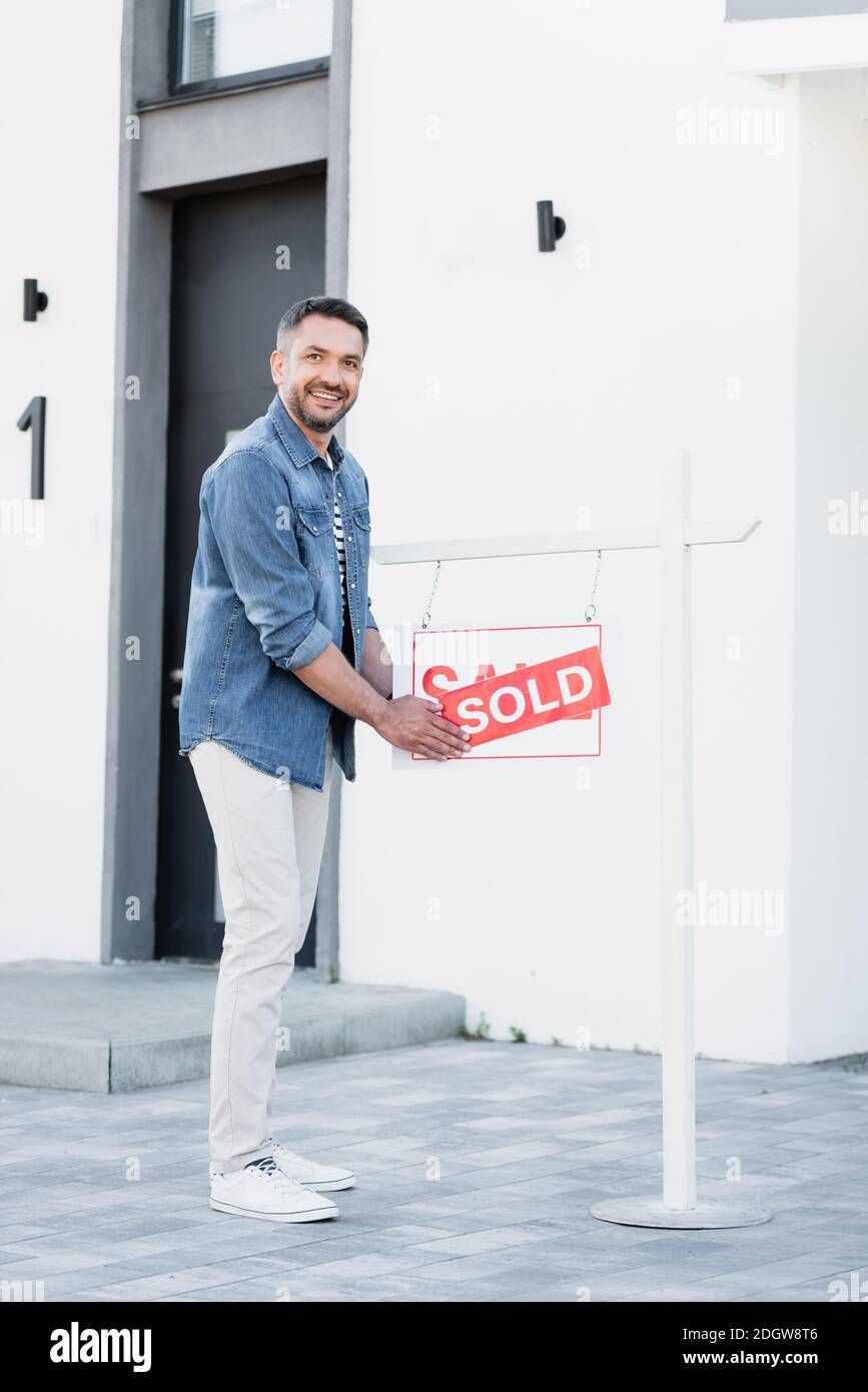 Full length of smiling man holding sign with sold lettering near house ...