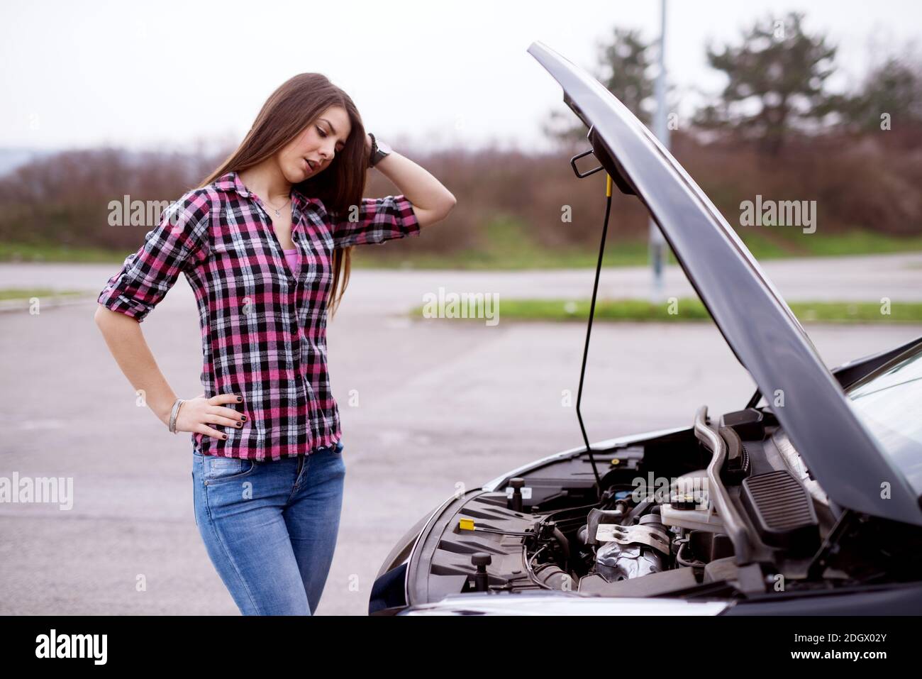 Side view of a young worried beautiful girl is looking under the hood of her car and holding her ...