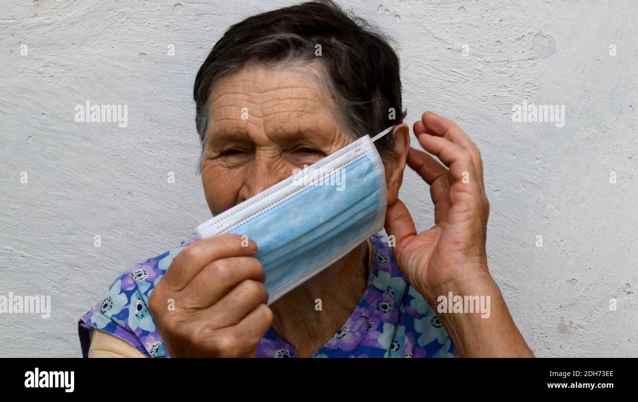 Senior wrinkled woman puts on protective medical mask touching her ear ...
