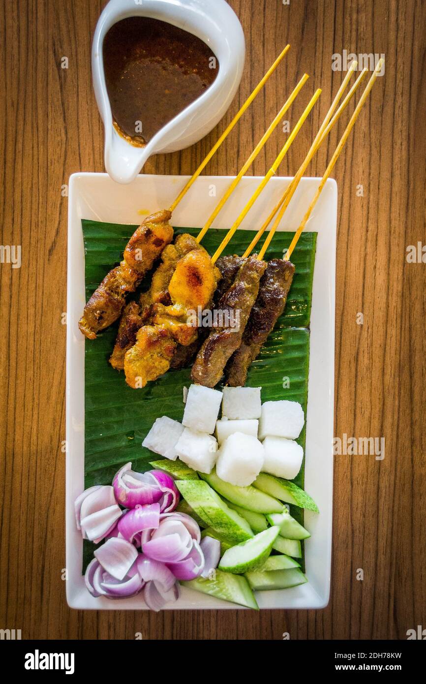 a plate of chicken and beef satay with raw onions, cucumber, rice cake ...