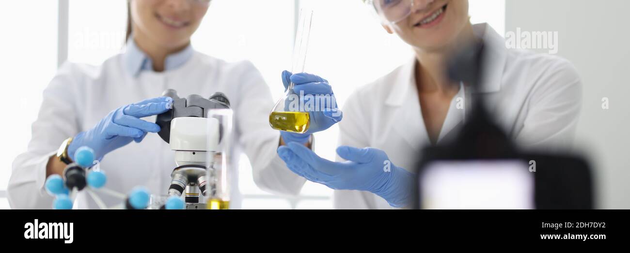 Two women in uniform and chemical goggles hold glass flask of yellow ...