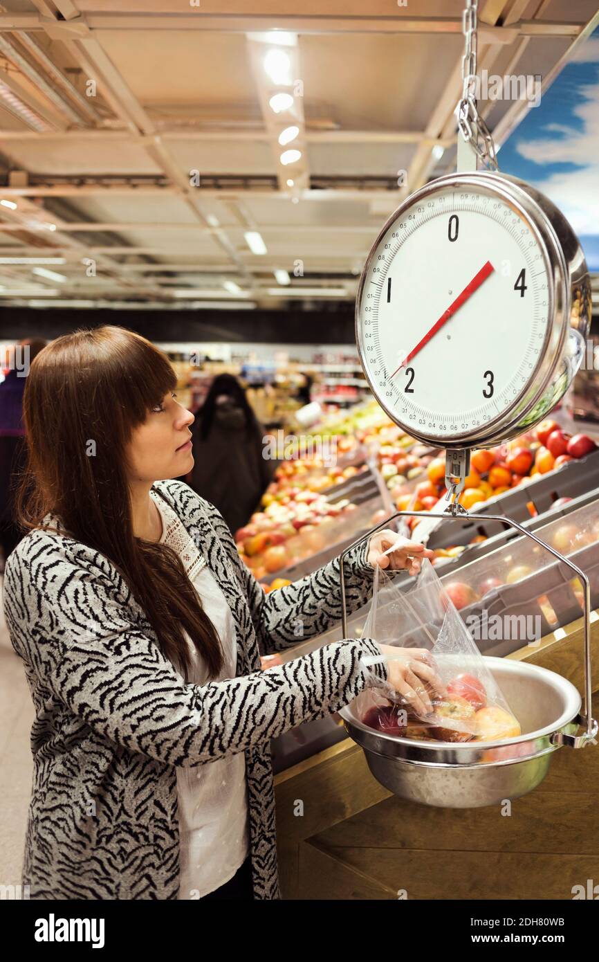 Woman looking at weight scale while weighing apples at supermarket ...