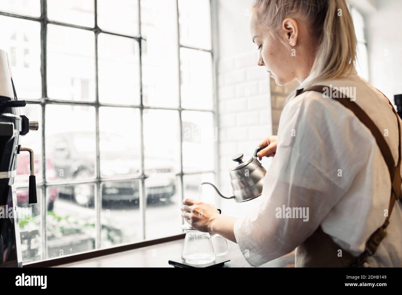 Side view of female barista preparing coffee at cafe counter by window ...