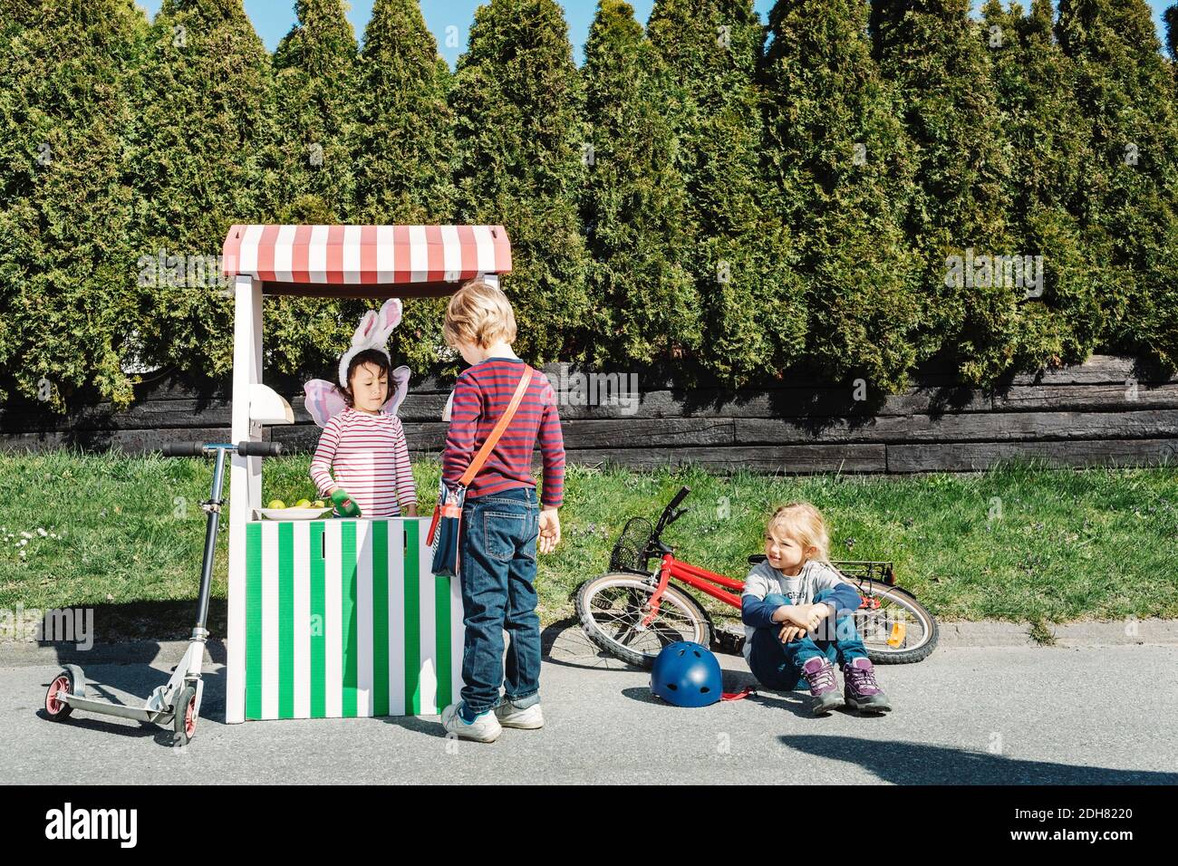 Boy standing at food stall while friend imitating as seller on footpath ...