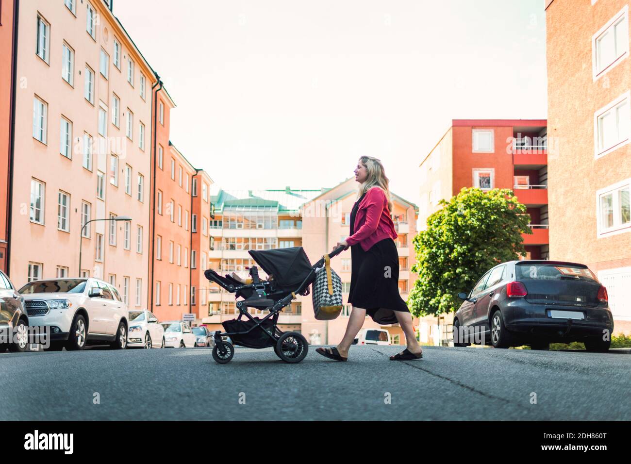 Full length side view of woman pushing baby in carriage crossing city ...
