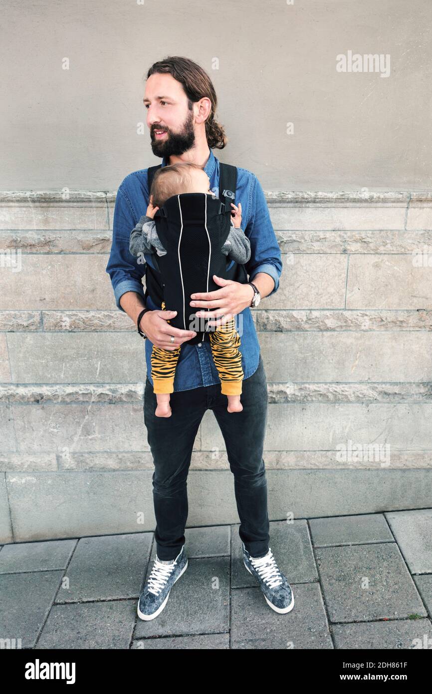 Full length of mid adult father carrying baby in carrier on sidewalk ...