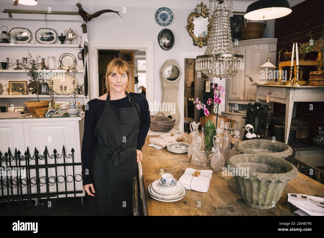 Portrait of confident female owner standing by table in antique store ...