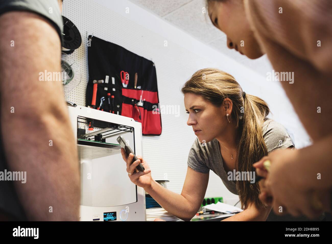 Female engineer holding smart phone and looking at 3D printer with ...