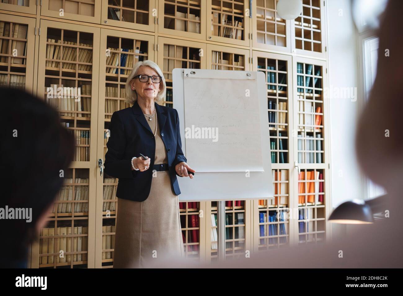 Senior woman giving presentation while standing against bookshelf Stock ...