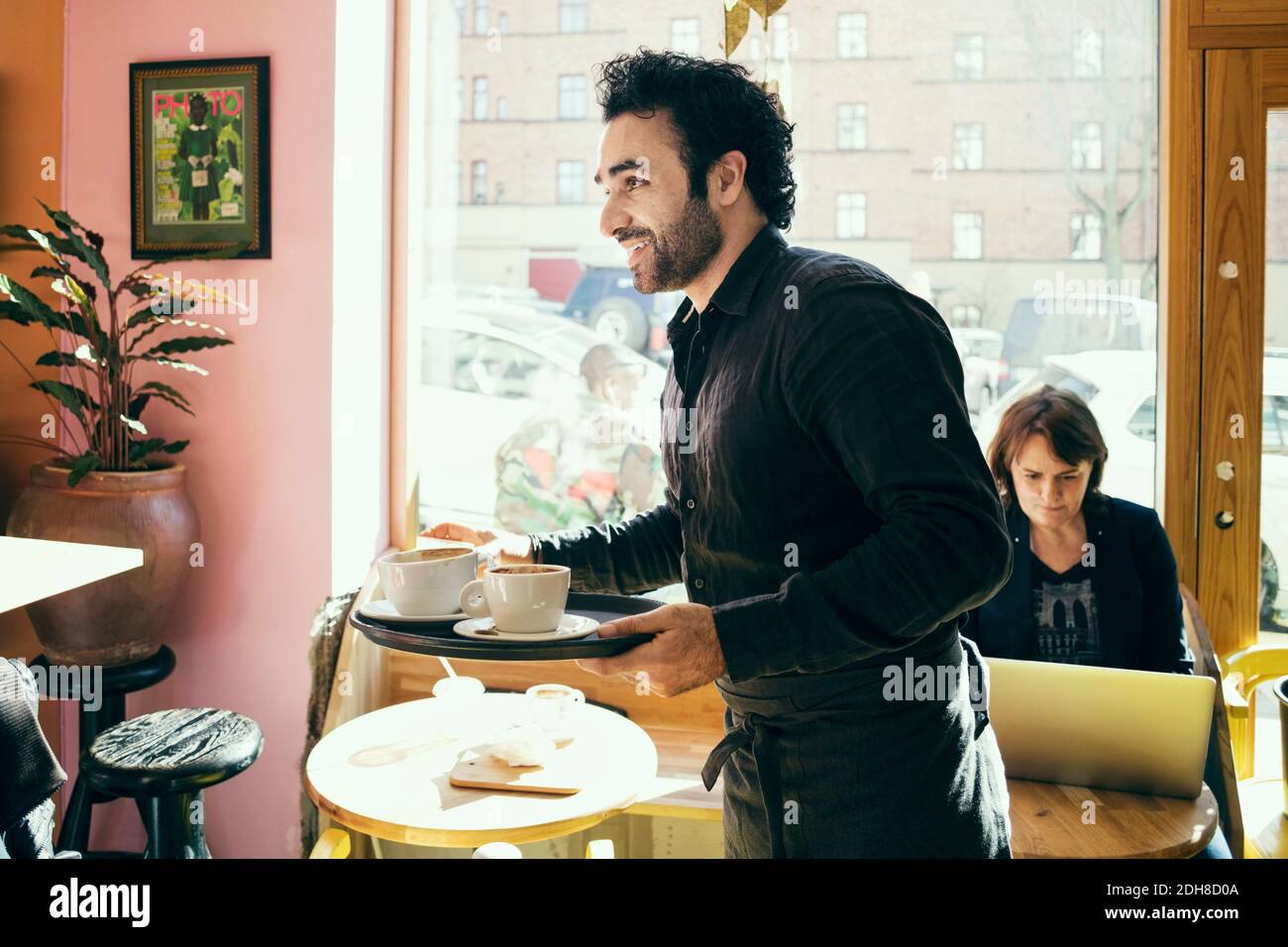Smiling waiter holding serving tray against customer using laptop in ...