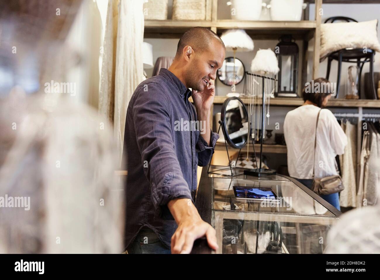 Smiling owner using phone while female customer looking at clothes in ...