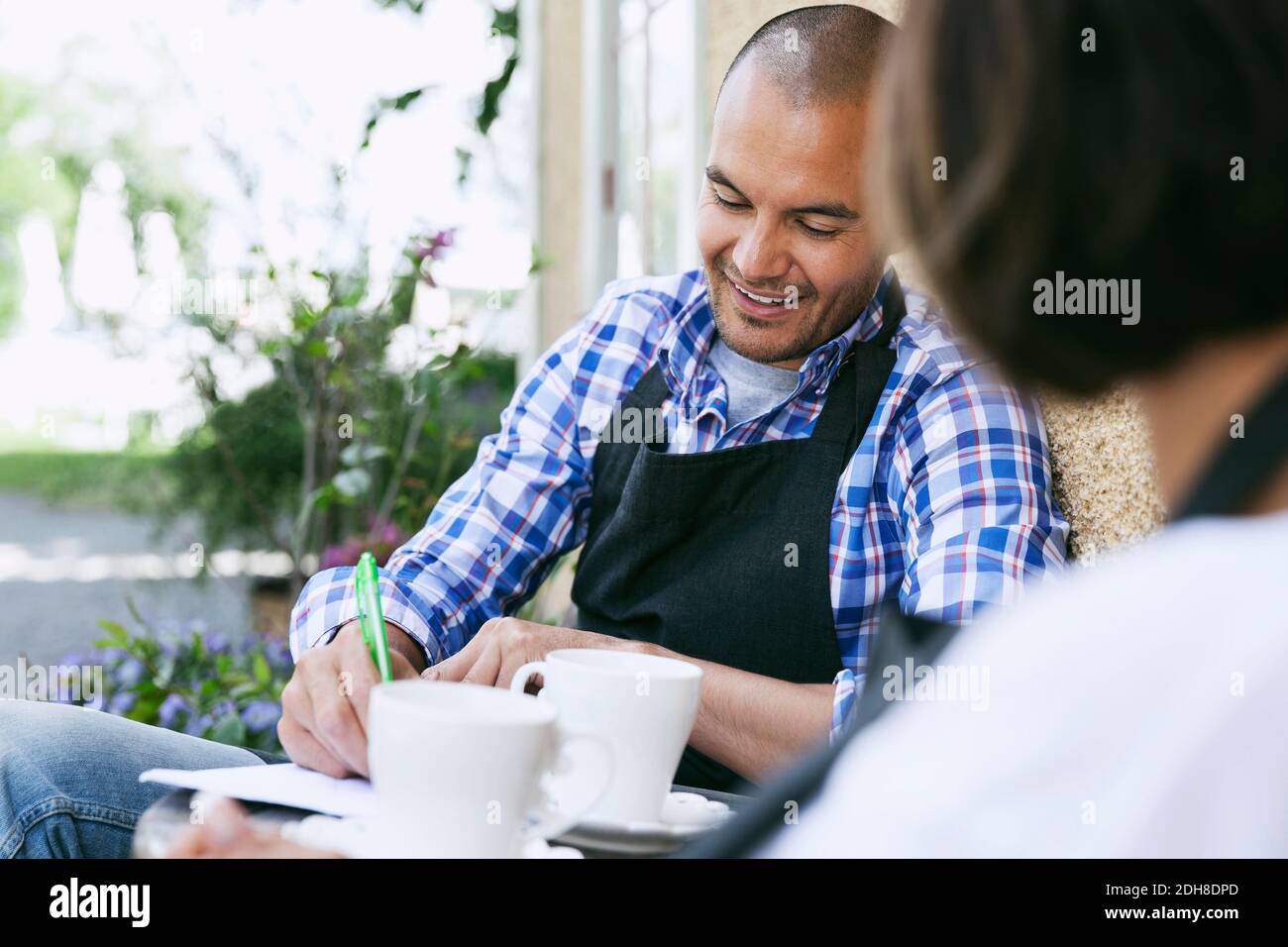 Smiling man writing on paper while having coffee with colleague outside ...