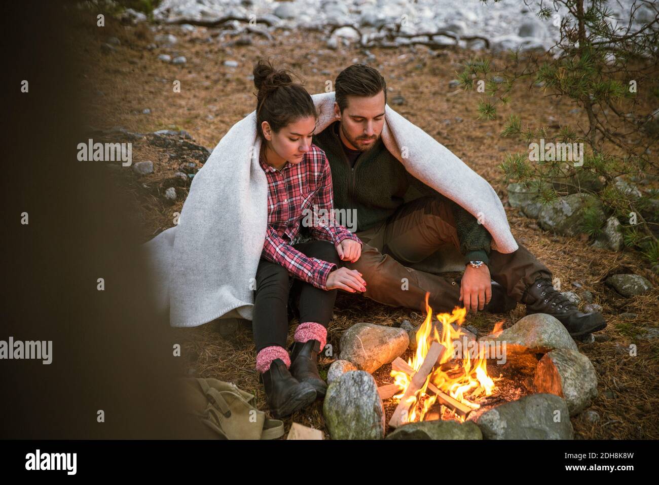Couple wrapped in blanket while sitting by fire pit at campsite Stock ...