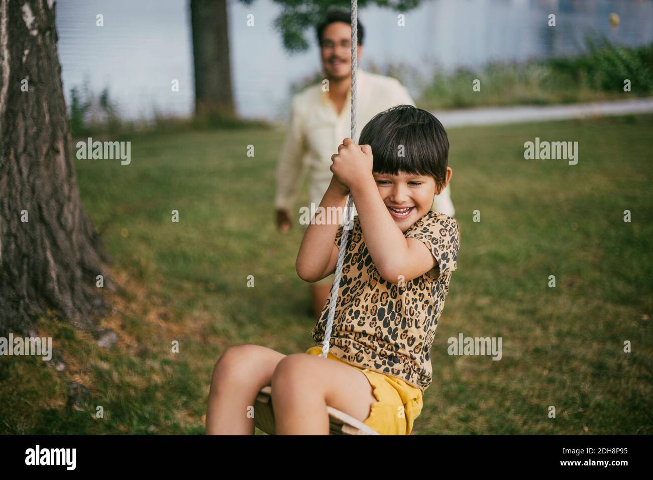 Smiling daughter sitting on rope swing while father standing in ...