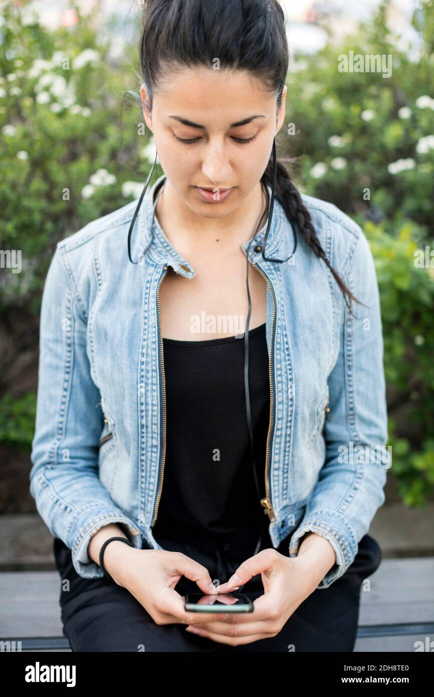 Young woman sitting on retaining wall while using phone in city Stock ...