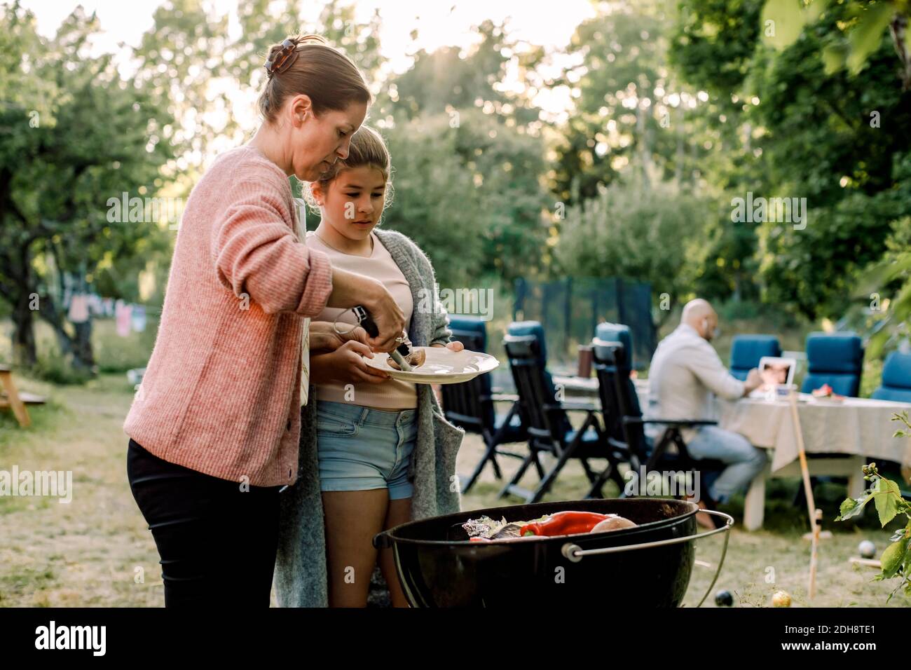 Side view of mother and daughter preparing food over barbecue grill in ...