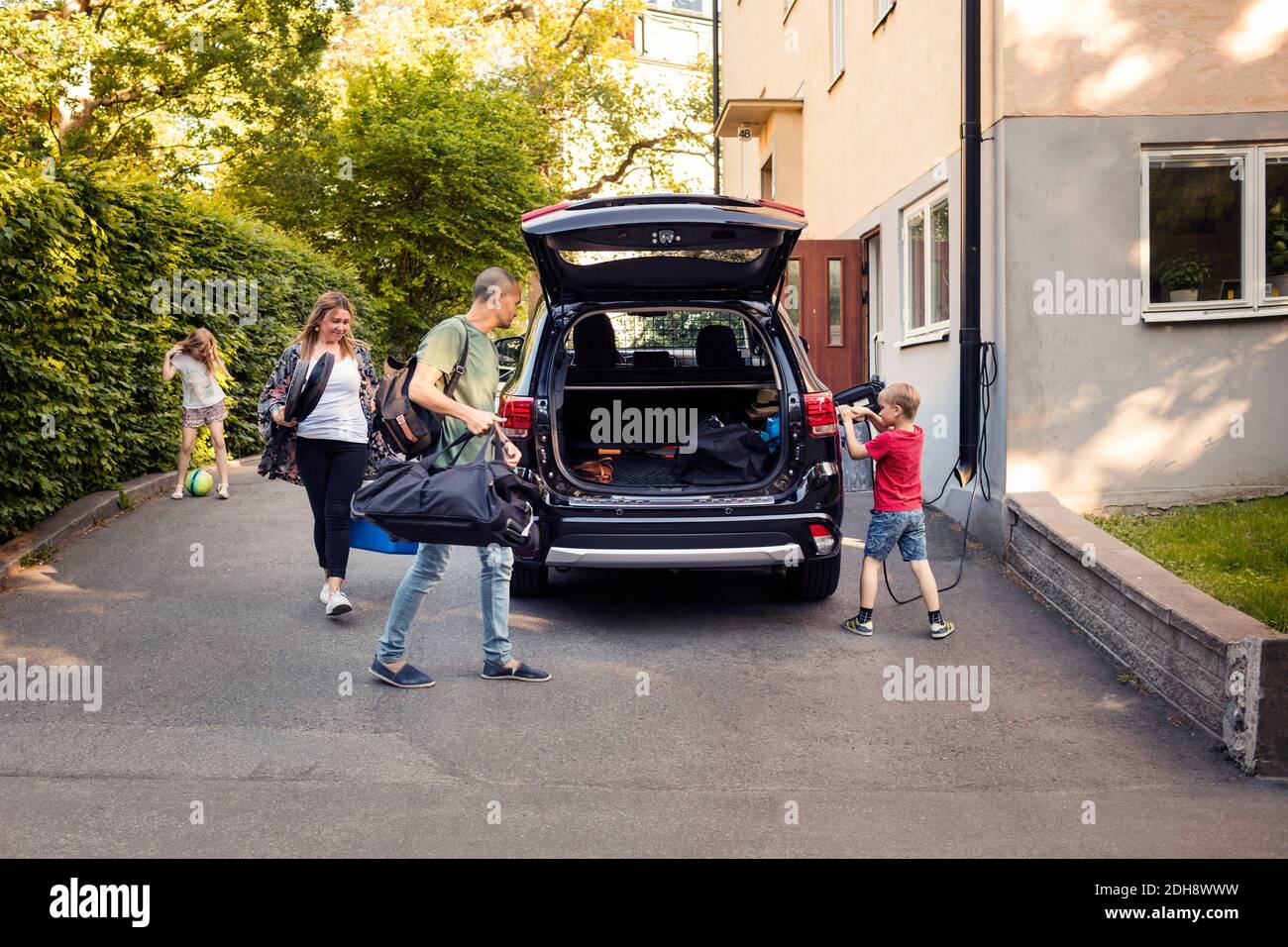 Man and woman loading luggage in trunk while boy charging electric car ...
