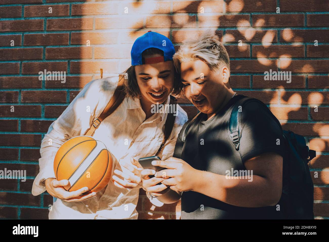 Smiling male friends using smart phone while standing against brick ...