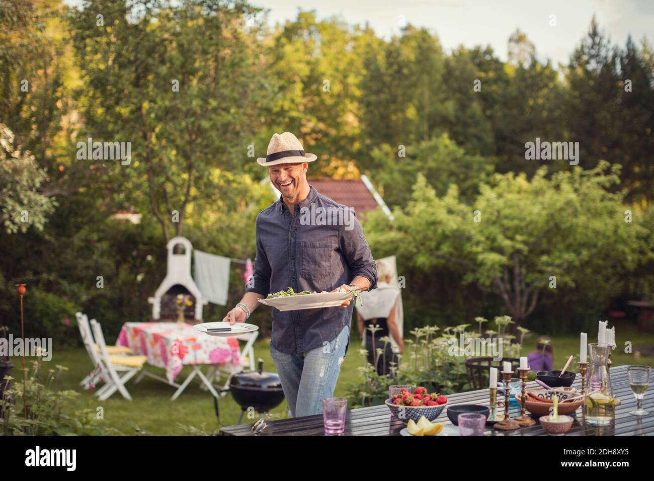 Cheerful man carrying plates by dining table in back yard during garden ...