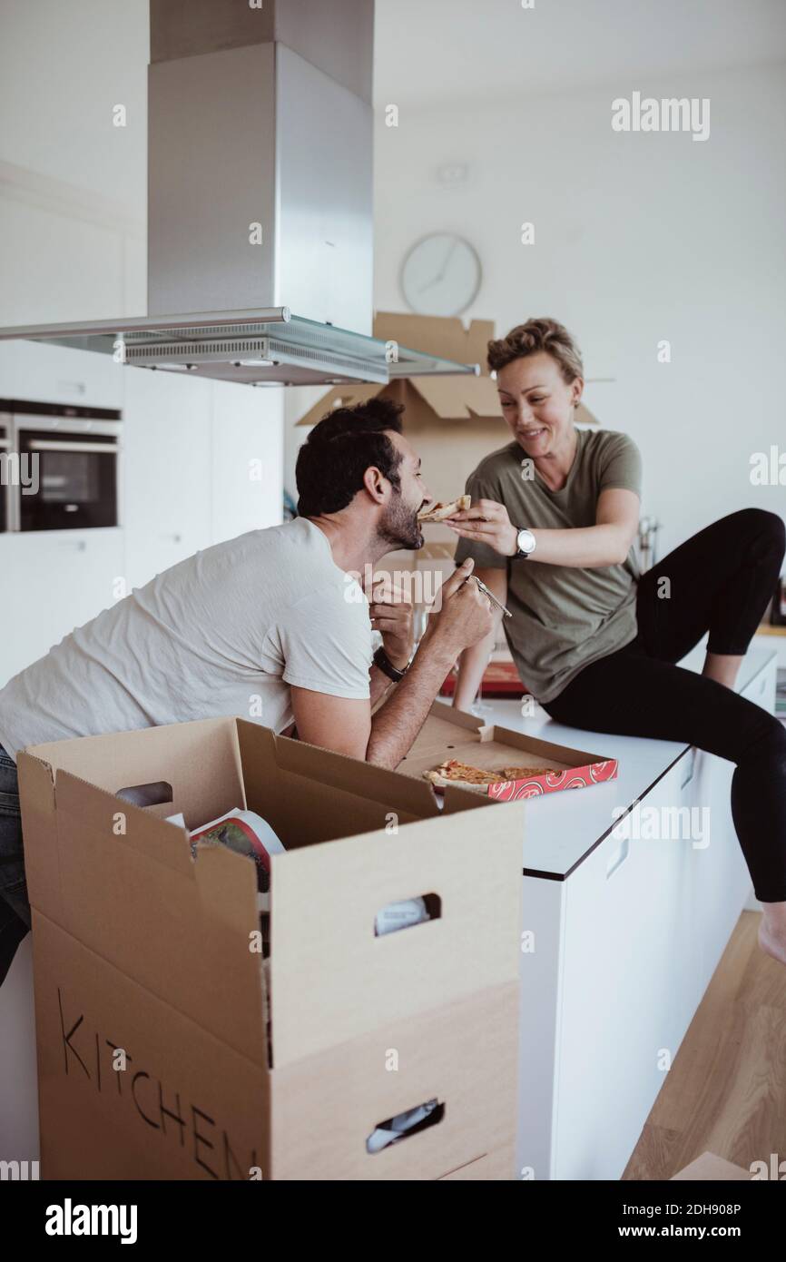 Smiling woman feeding pizza to male partner while sitting on kitchen ...
