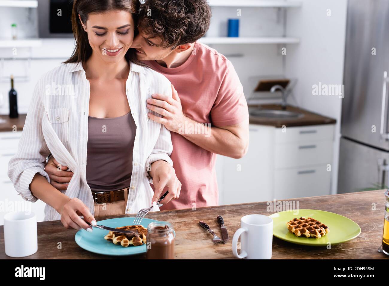 Young man hugging smiling girlfriend near waffles and cups on kitchen ...