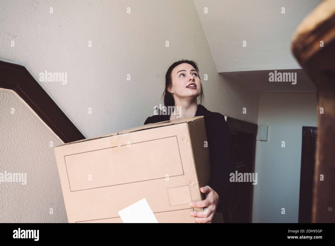 Confident delivery woman with cardboard box looking up against wall ...