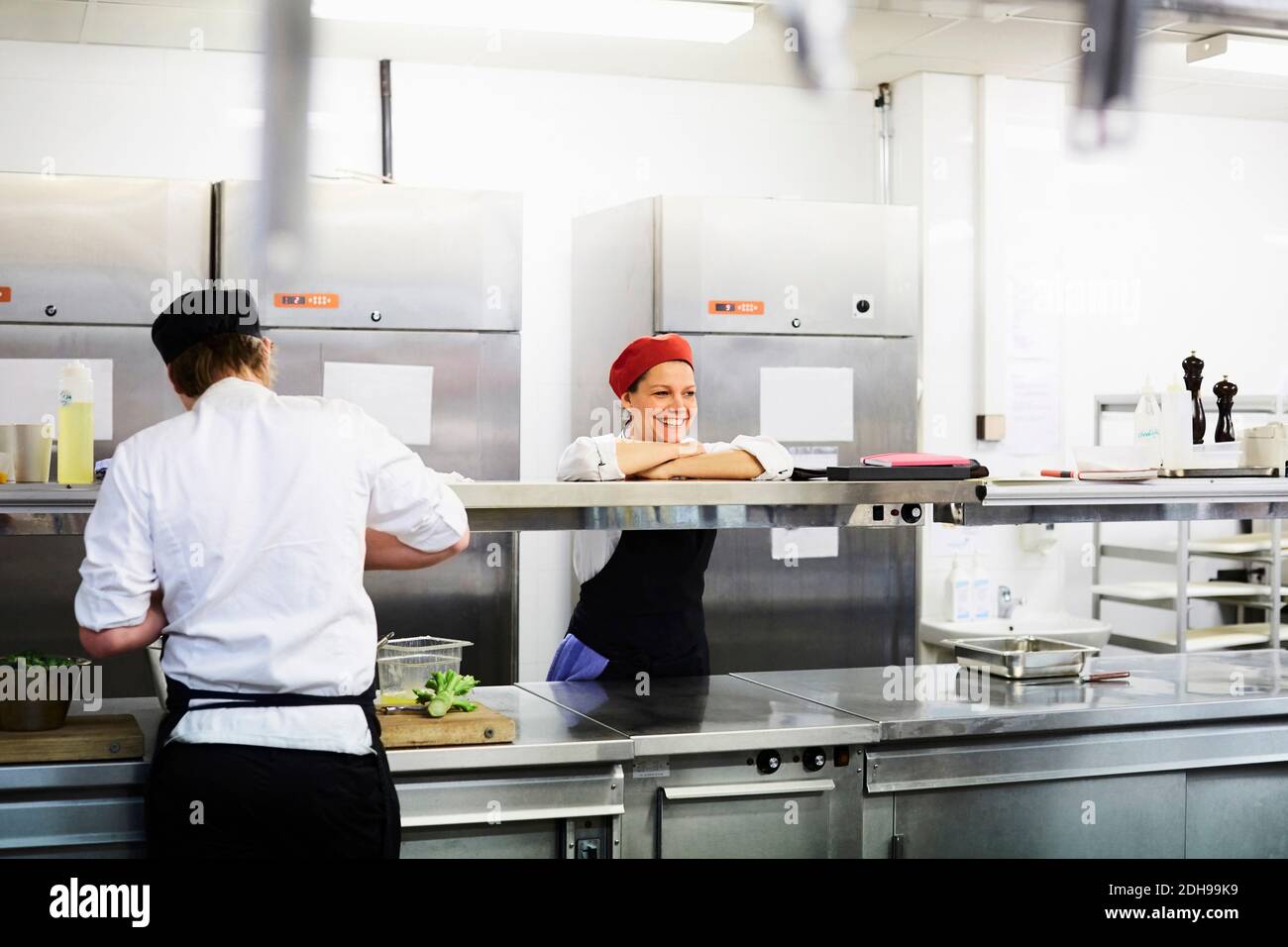 Smiling female chef leaning on counter with student working in cooking ...