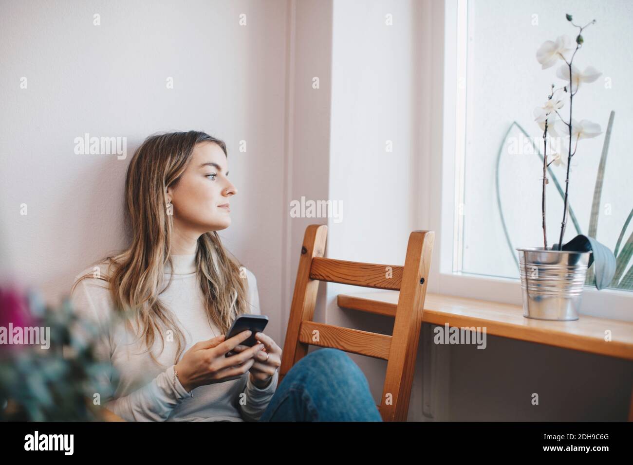 Young woman sitting with mobile phone on chair while looking through ...
