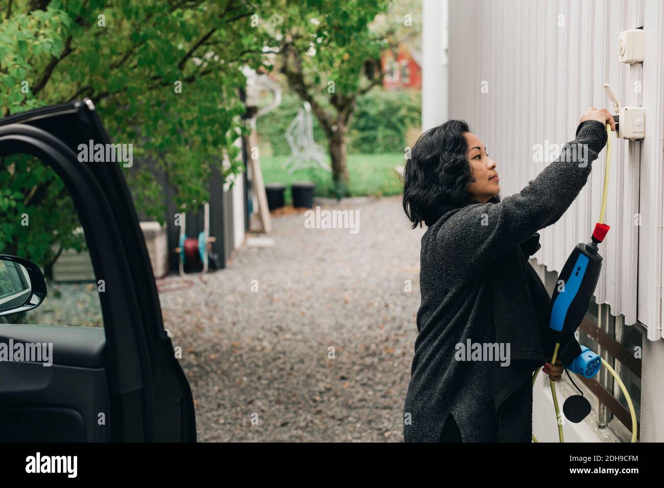 Side view of woman connecting plug to outlet for charging electric car ...