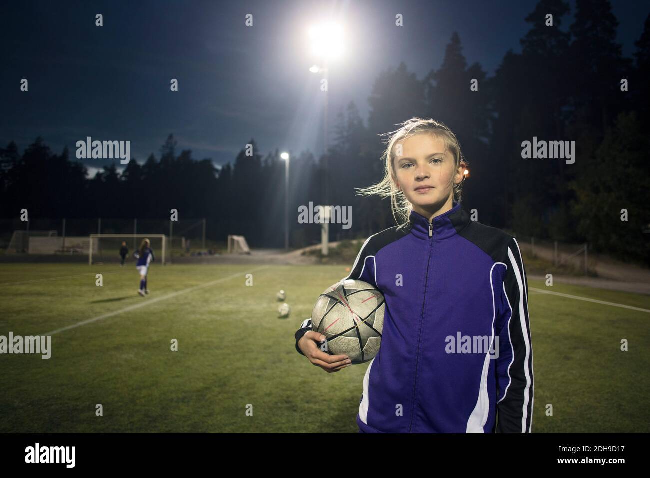 Portrait of girl standing with soccer ball on field against trees at ...