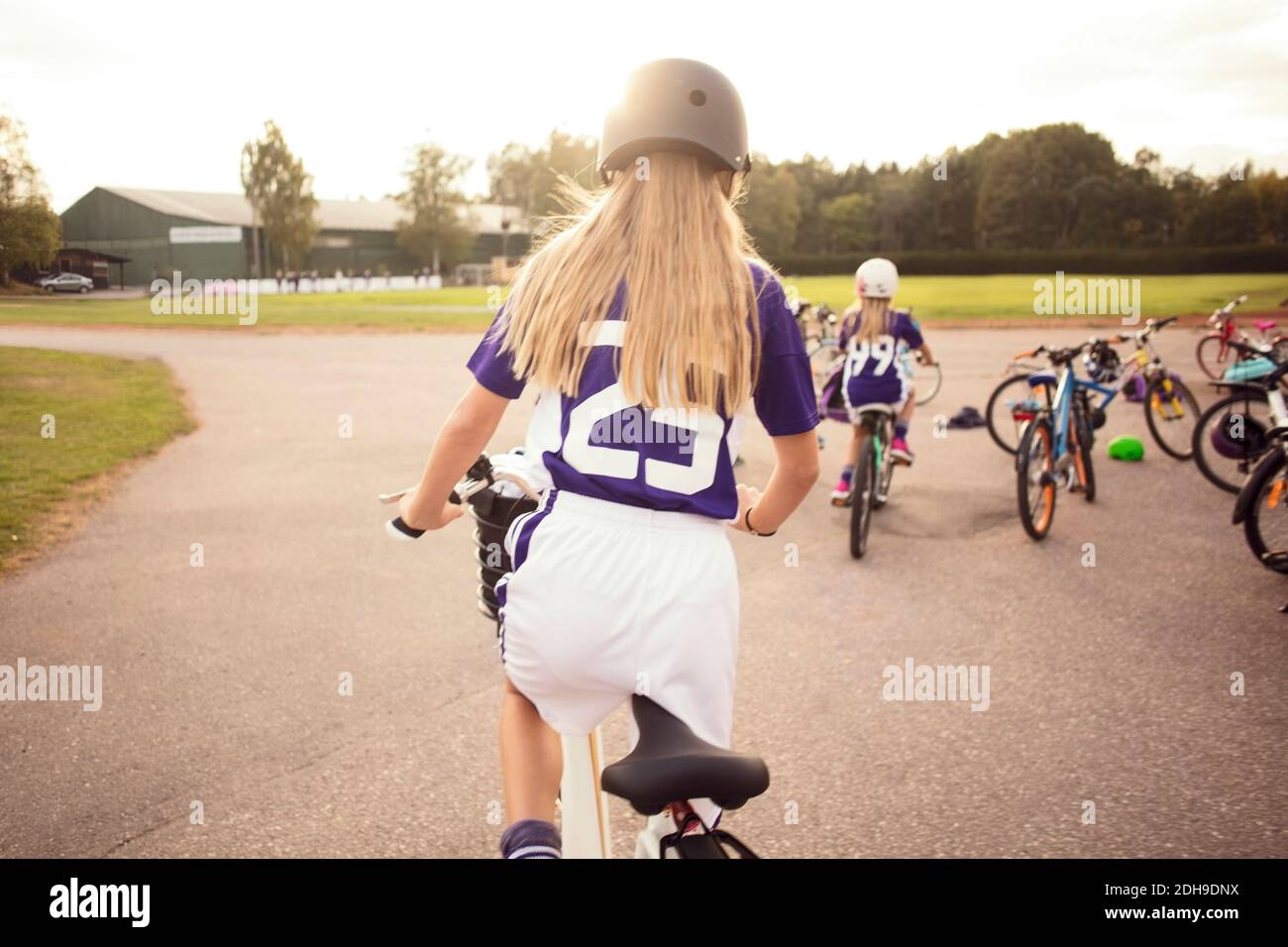 Rear view of girls cycling on footpath by soccer field against sky ...