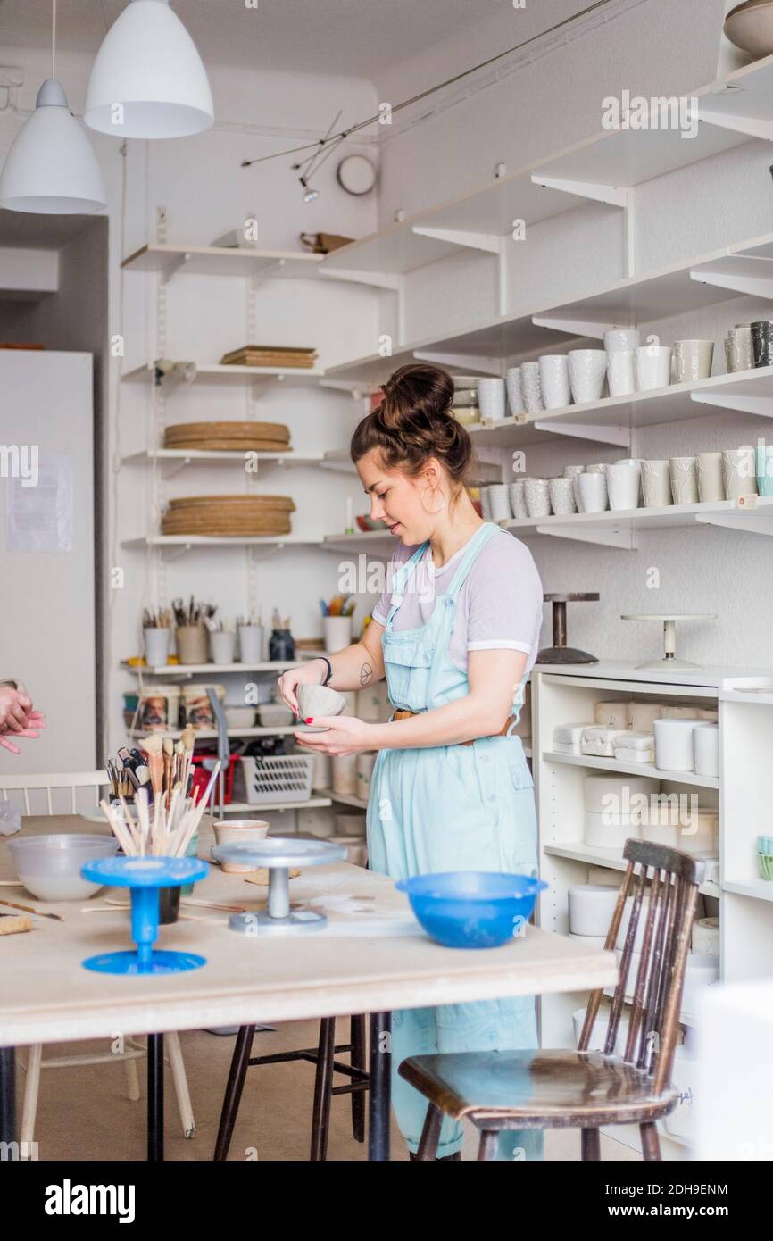 Young female potter molding shape from clay while standing by workbench ...