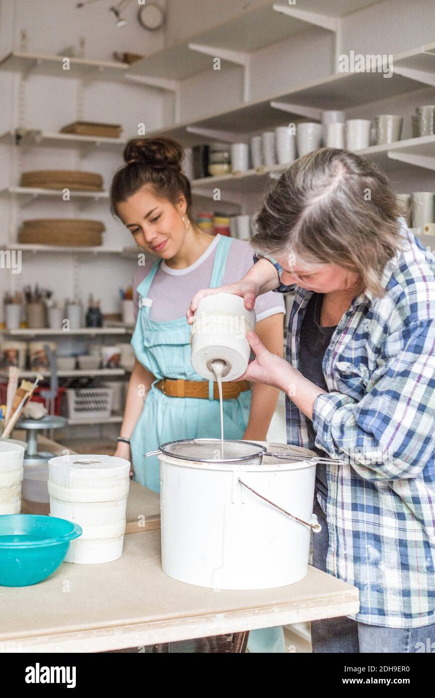 Young employee looking at mature female potter pouring clay from vase ...