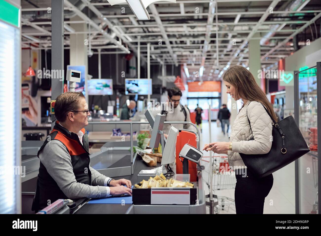 Mature cashier looking at woman paying through technology at checkout ...