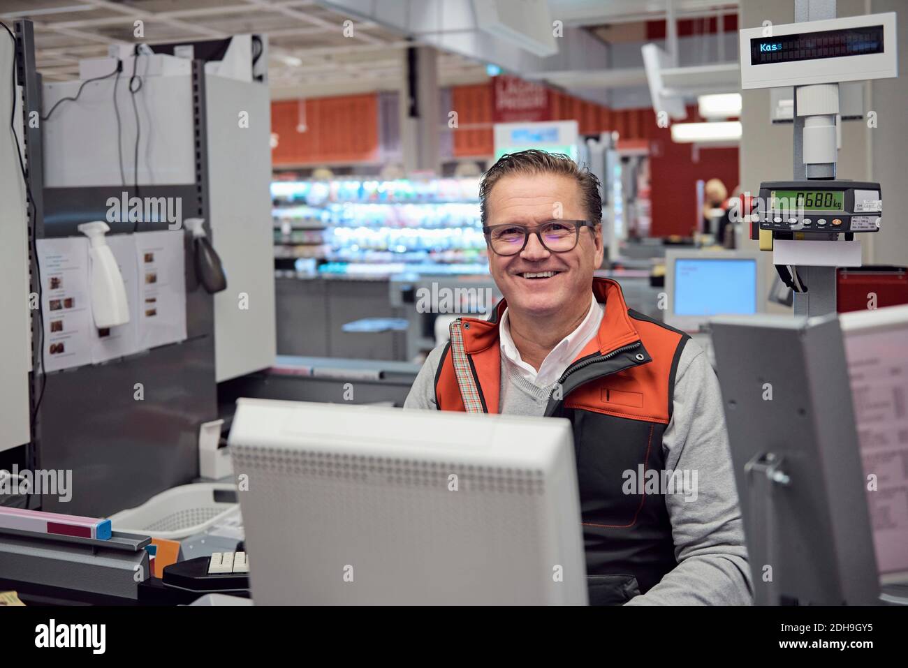 Portrait of smiling mature cashier sitting at checkout counter in ...