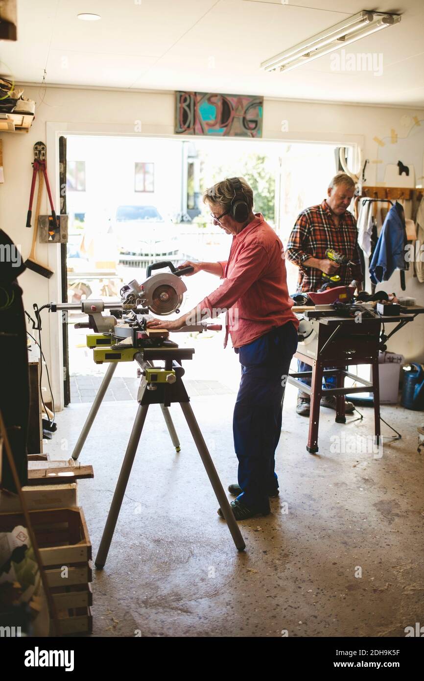 Side view of senior woman using circular saw while man standing in ...