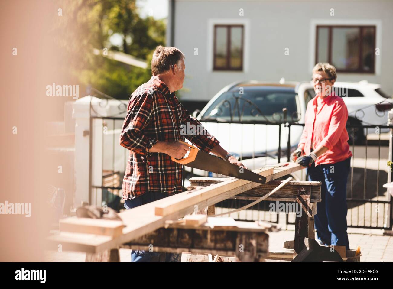 Senior man holding hand saw while looking at woman by wooden planks in ...