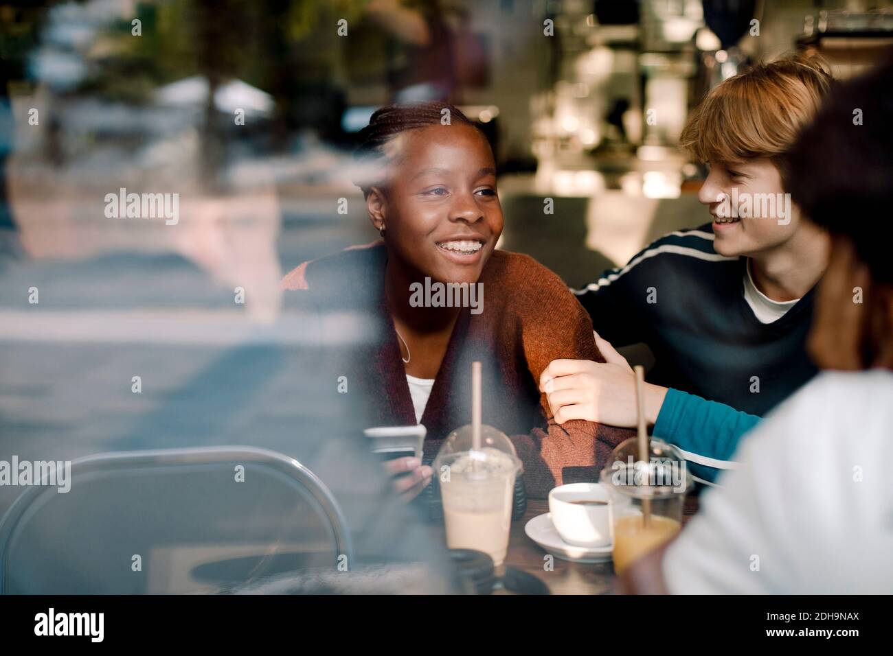 Smiling teenage girl with friends at cafe seen through glass window ...